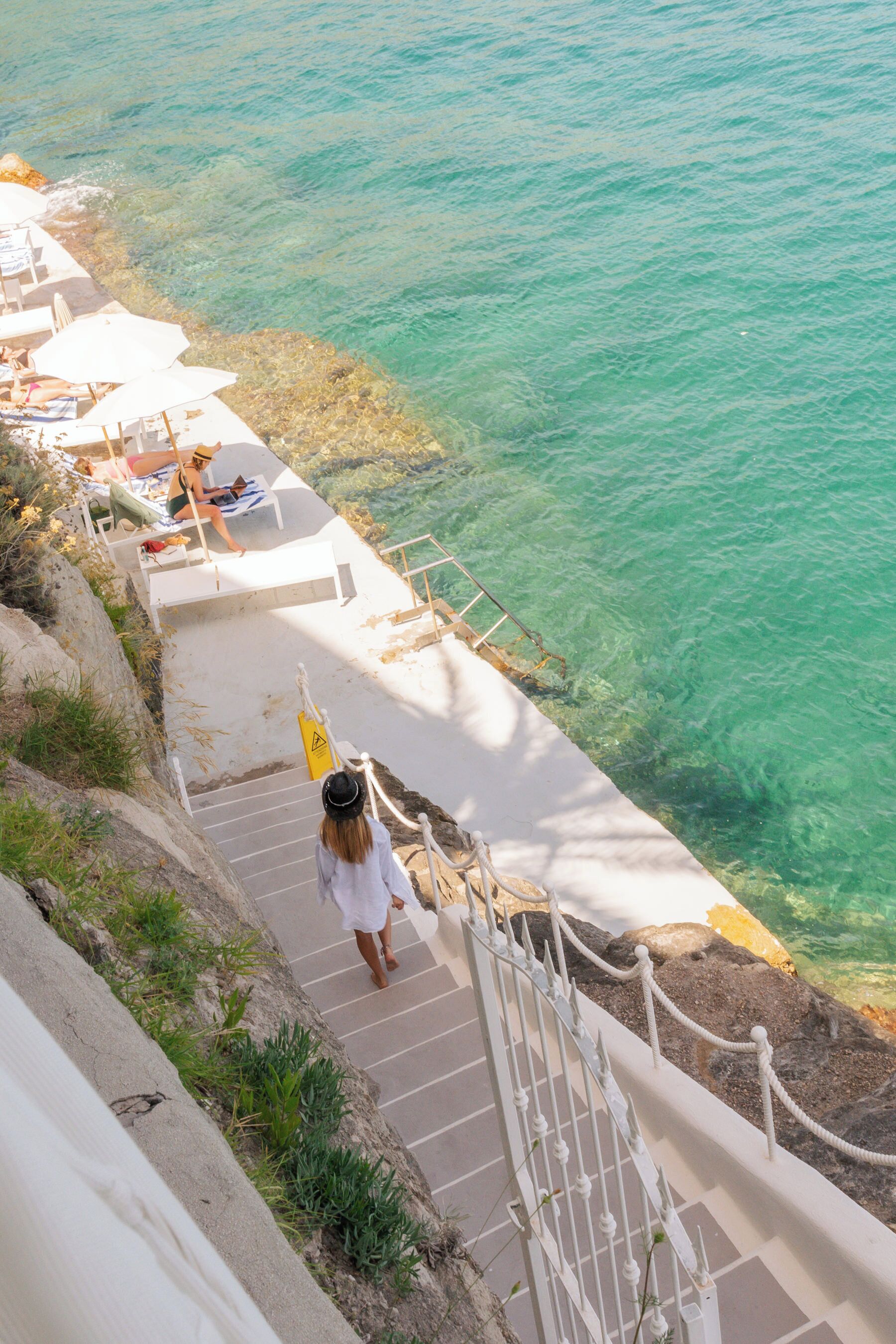 Escadaria que dá acesso a uma área de praia com água cristalina, cadeiras de praia e guarda-sóis.