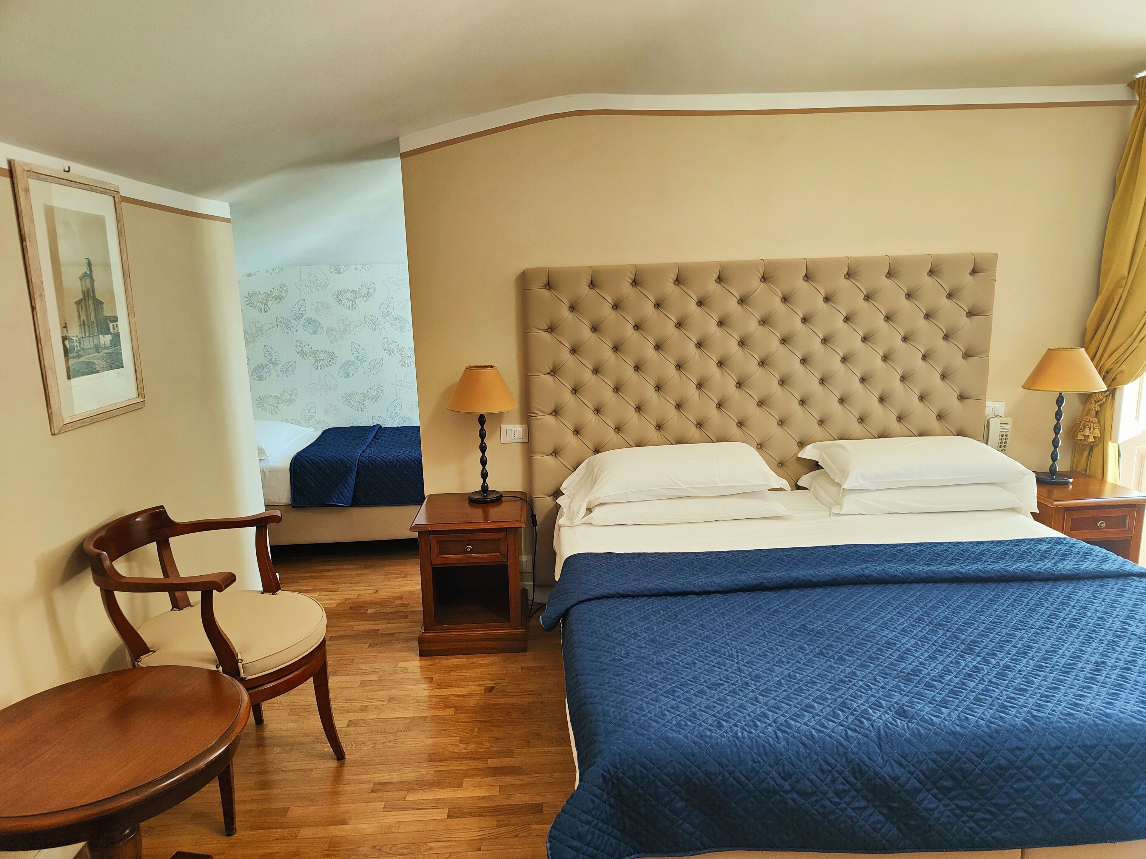 Quarto de hotel com uma cama grande, uma cadeira de madeira, uma mesa de cabeceira com abajur e uma parede com um quadro emoldurado.