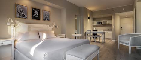 Quarto de hotel moderno com uma cama grande, uma mesa pequena, uma cadeira e uma kitchenette ao fundo.