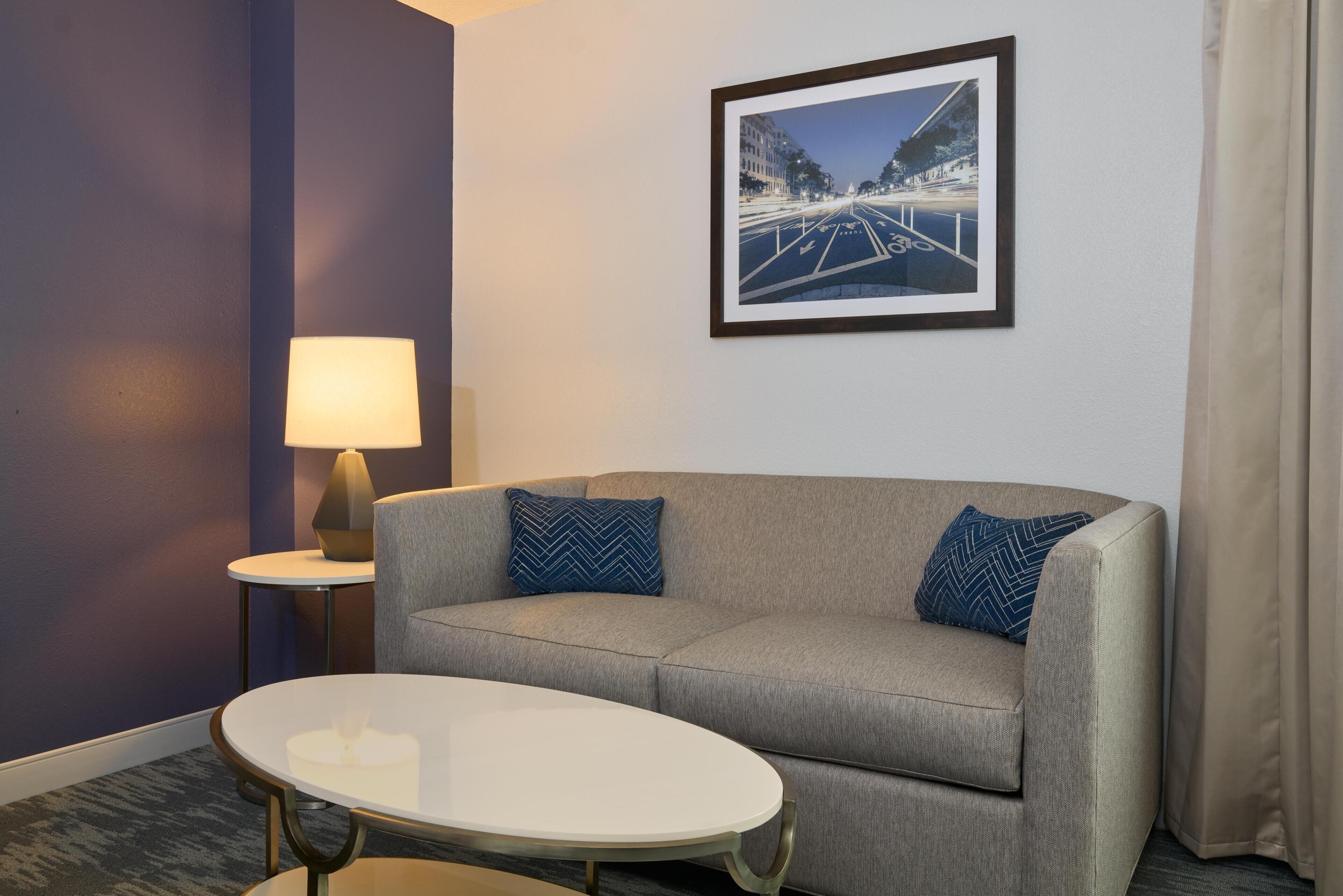 Quarto de hotel com sofá cinzento, uma pequena mesa redonda, luminária e quadro pendurado na parede.