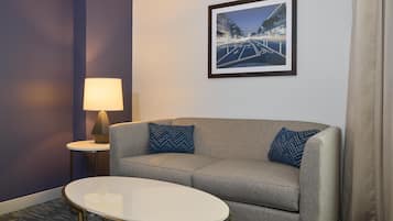 Quarto de hotel com sofá cinzento, uma pequena mesa redonda, luminária e quadro pendurado na parede.
