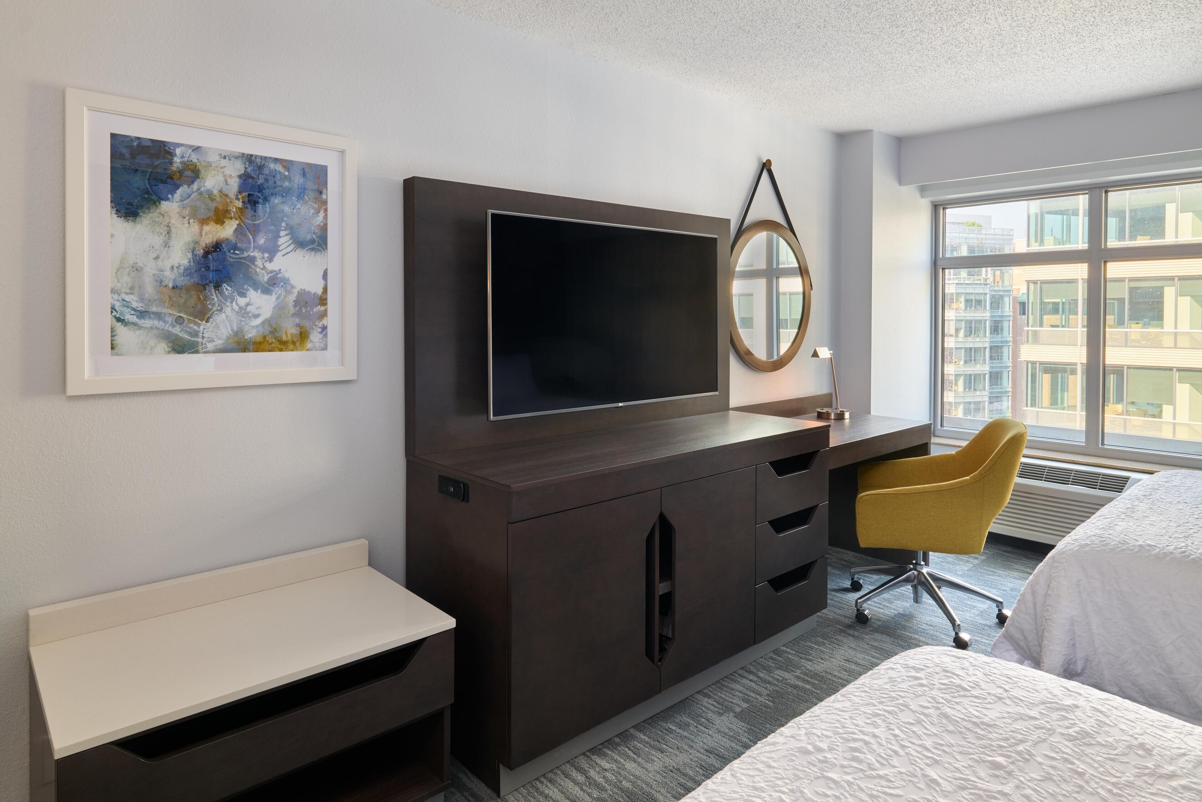 Quarto de hotel moderno com televisão de tela plana grande, escrivaninha com cadeira, cama e janela ampla com vista para a cidade.