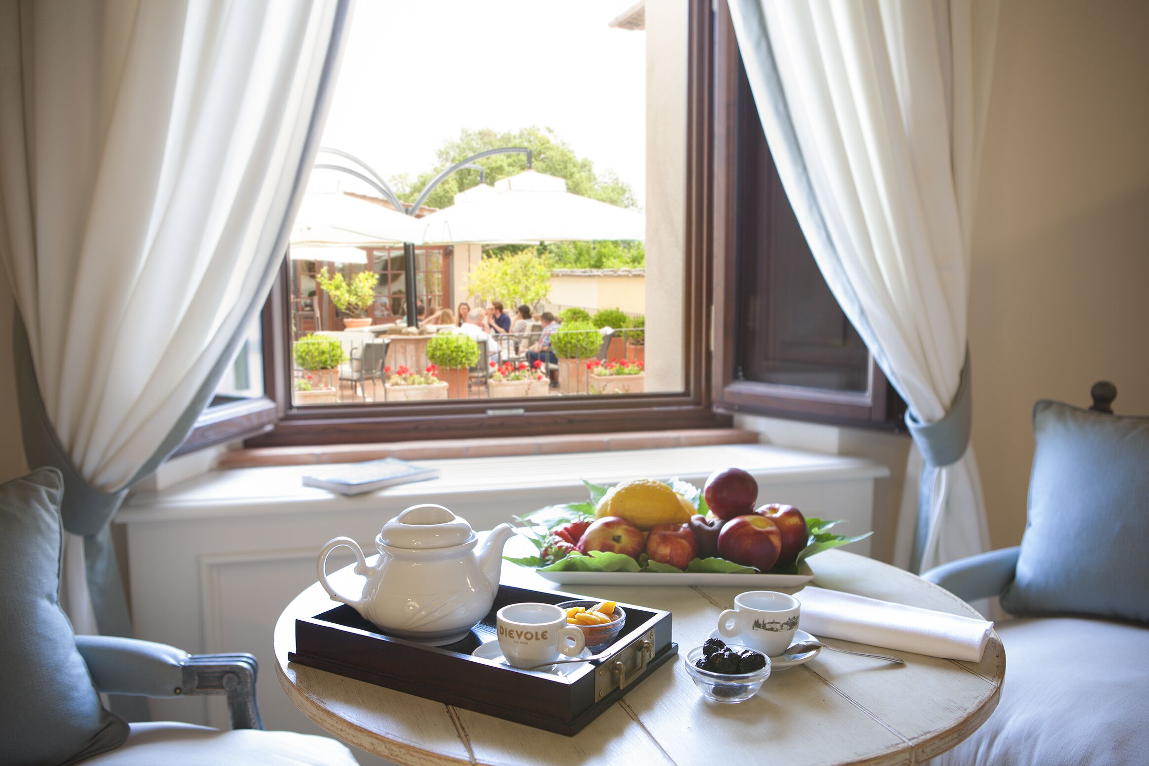 Bandeja de café da manhã com bule, xícara e frutas sobre uma mesa próxima a uma janela com cortinas.