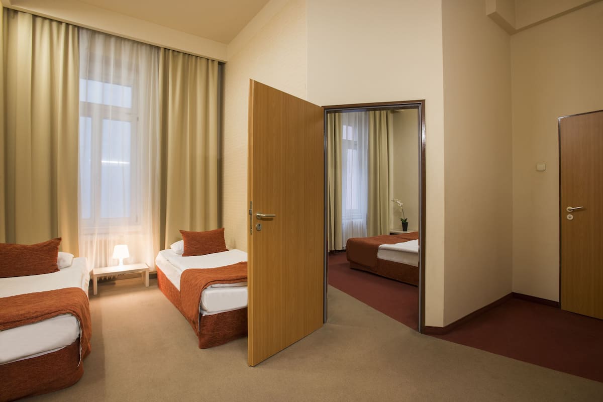 Quarto de hotel com duas camas, uma porta de madeira e vista pela janela com cortinas.