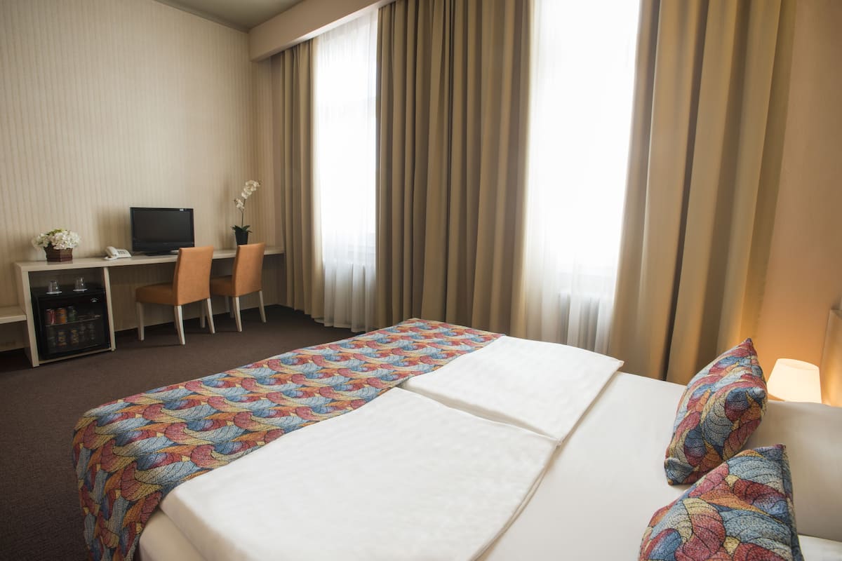 Quarto de hotel com uma cama, uma escrivaninha com computador, uma cadeira e uma janela com cortinas.