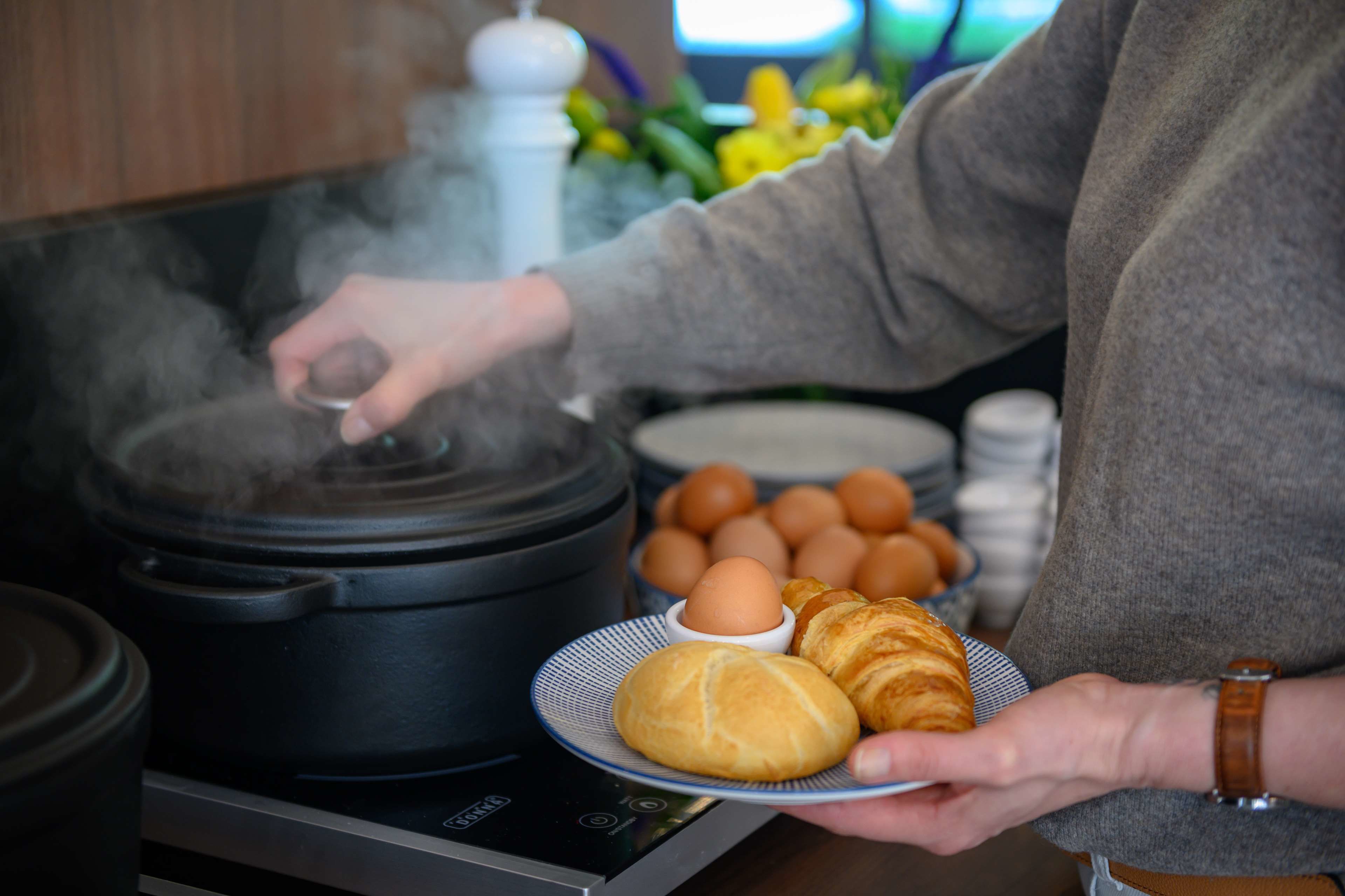 Uma pessoa segurando um prato com ovos e croissants, em pé perto de um fogão com uma panela soltando vapor.