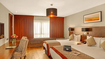 Quarto de hotel com secretária de madeira, duas camas, uma janela grande com cortinas, televisão fixada na parede e um quadro na parede.