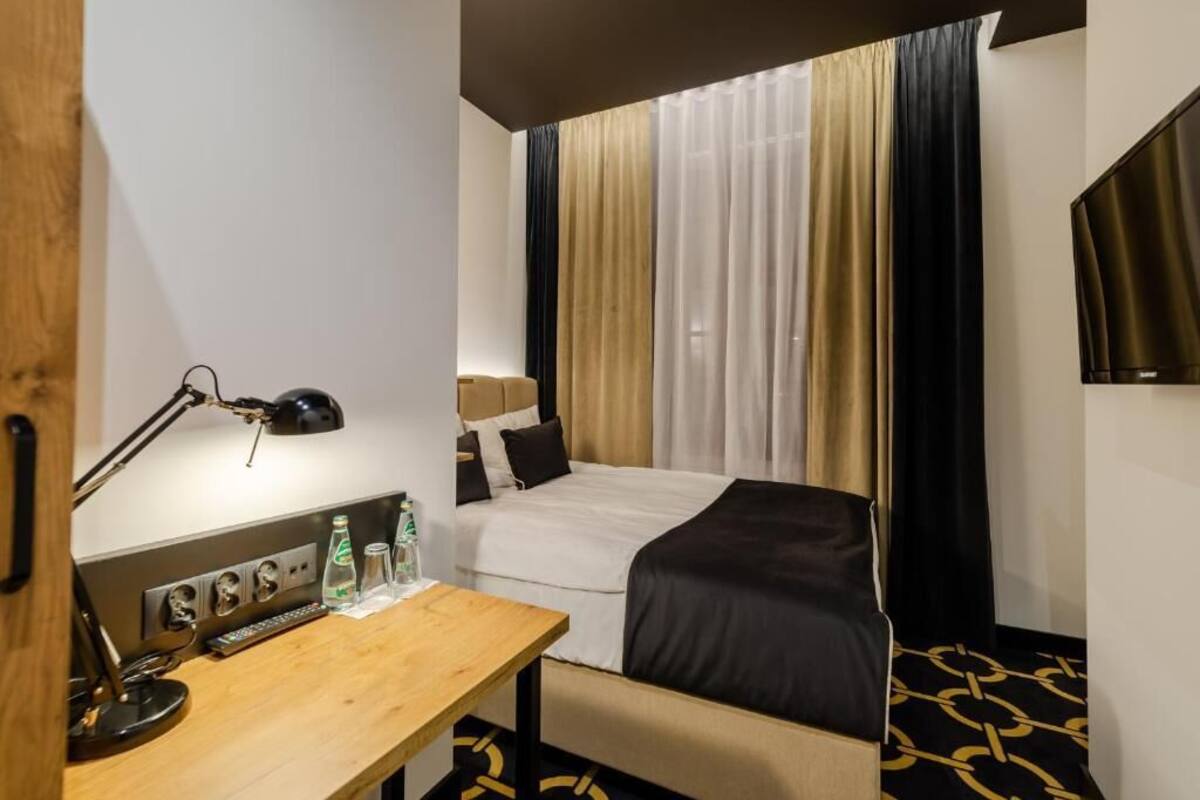 Quarto de hotel com uma cama, uma escrivaninha com luminária, uma televisão e uma janela com cortinas.
