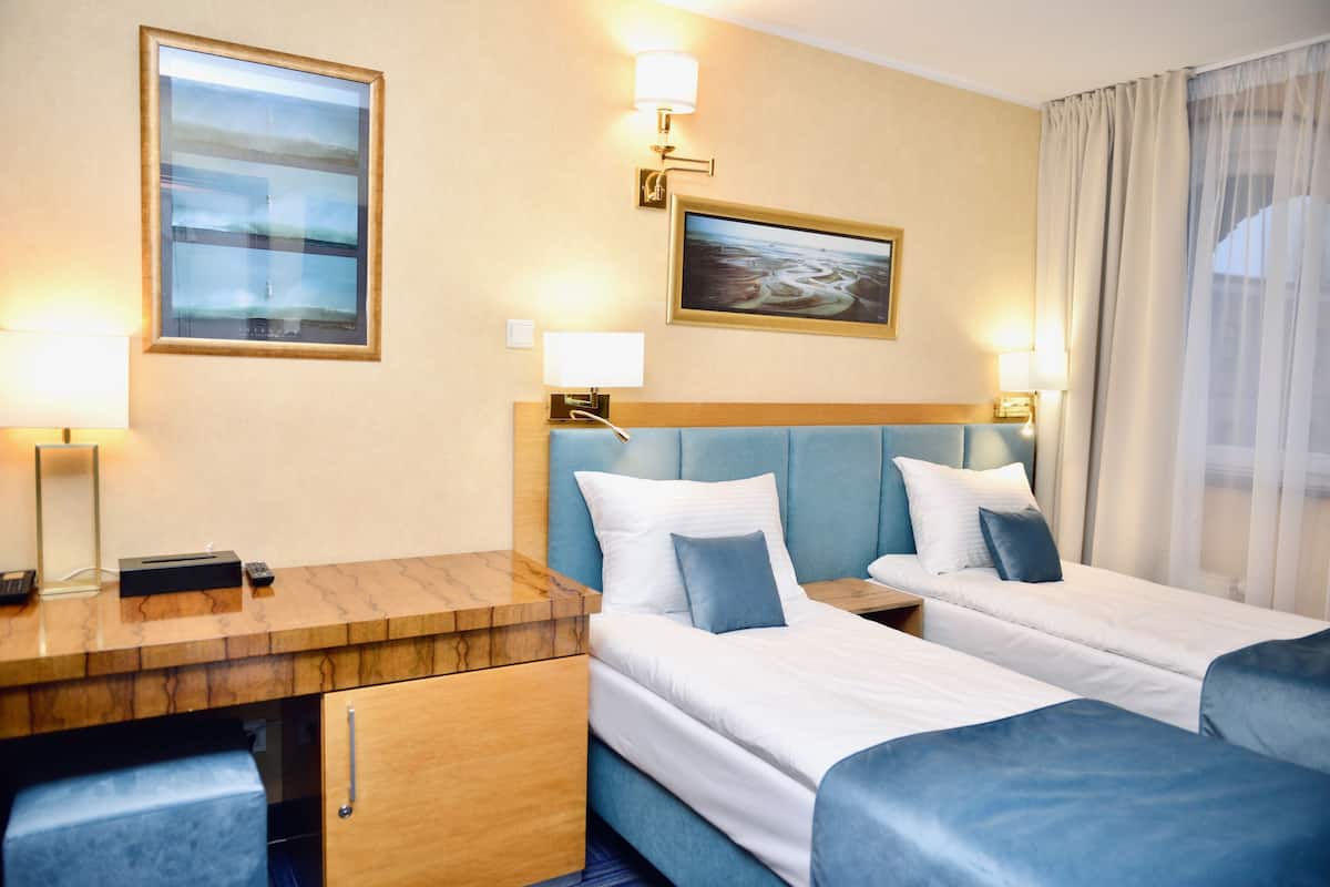 Quarto de hotel com duas camas, uma secretária de madeira, um minibar e uma janela grande com cortinas.