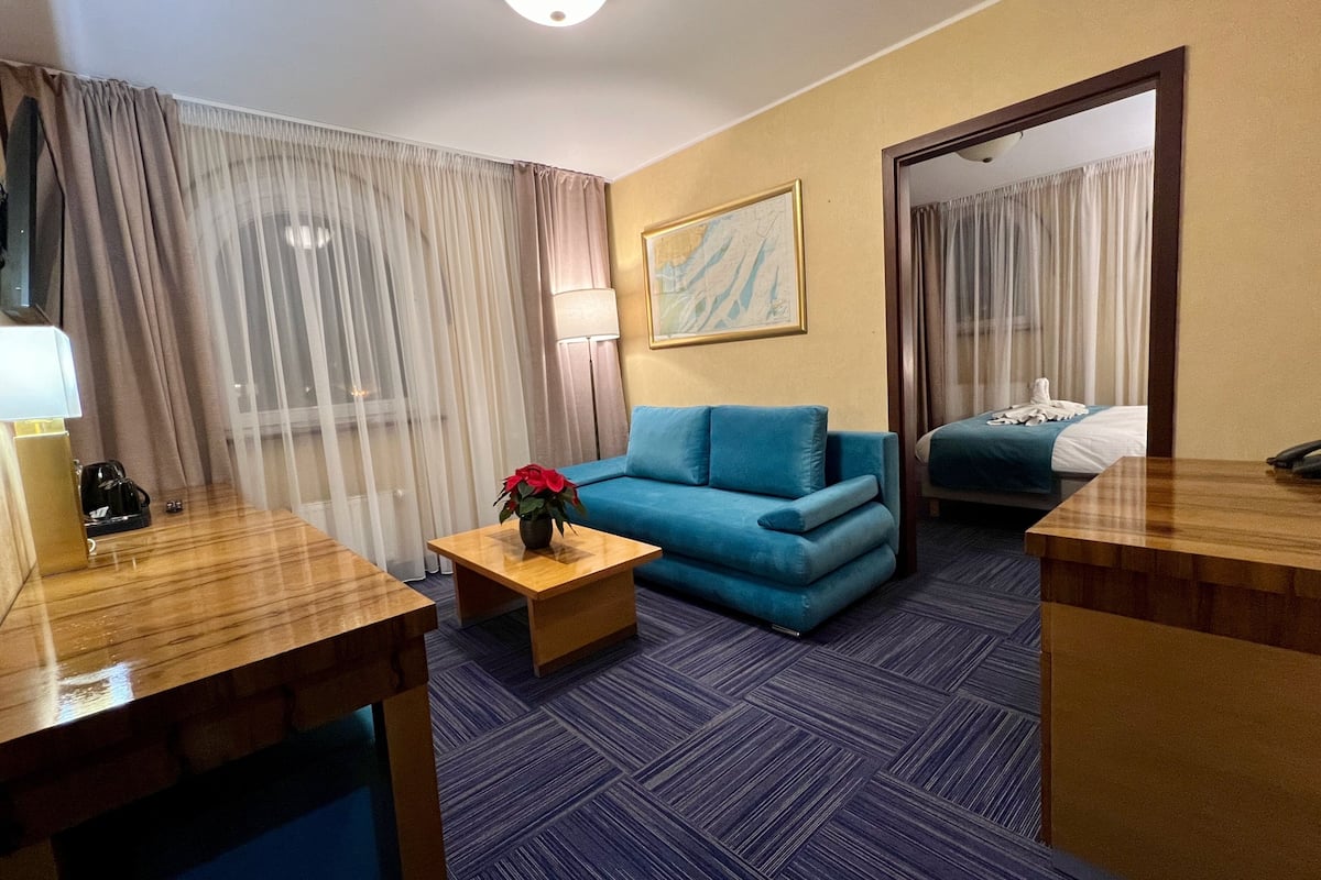 Quarto de hotel com um sofá azul, uma mesa de centro de madeira com um vaso de flores, uma escrivaninha com telefone e um espelho grande na parede.