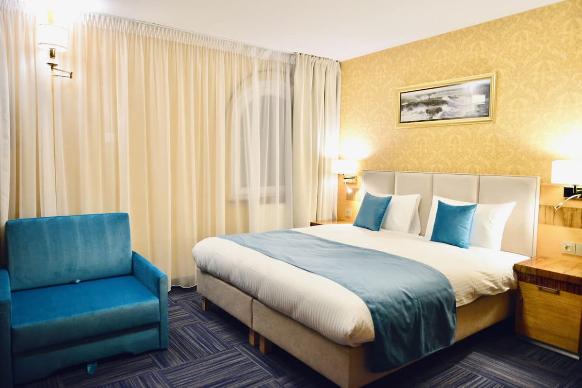 Quarto de hotel com uma cama grande, uma poltrona azul e uma mesa de cabeceira com abajur.