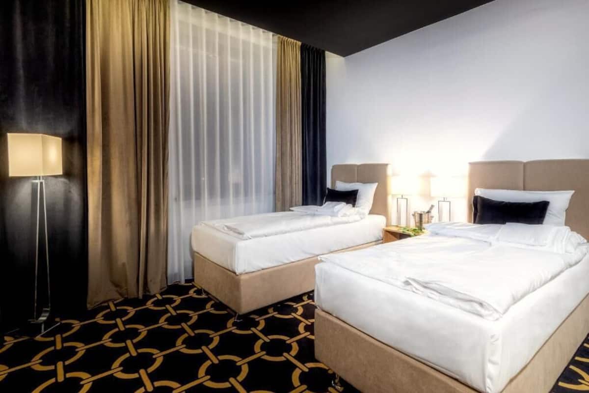 Quarto de hotel com duas camas, uma luminária de chão e uma janela grande com cortinas.