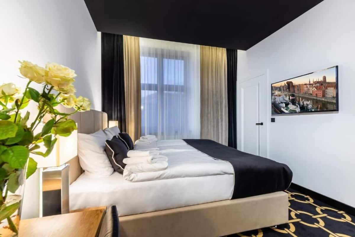 Quarto de hotel moderno com uma cama, mesa de cabeceira, abajur, janela com cortinas leves, quadro com vista urbana e um vaso de planta.