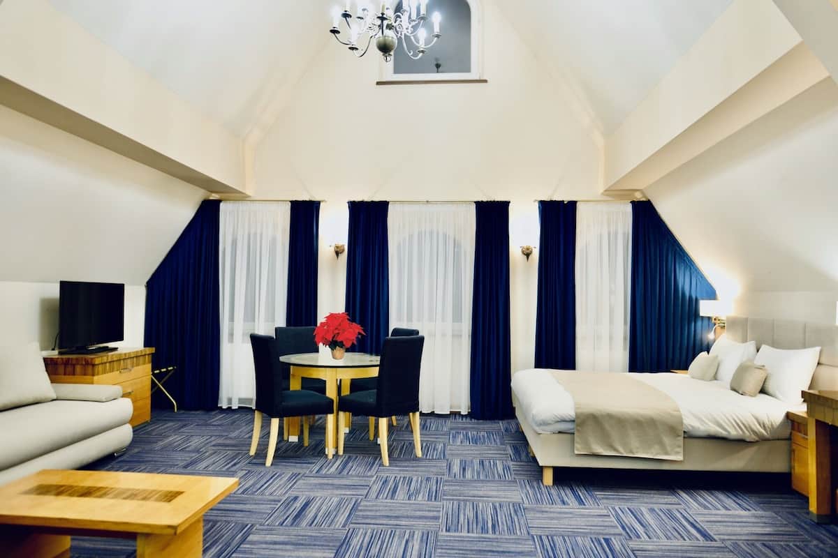 Quarto de hotel com cama, sofá, mesa de jantar com cadeiras, televisão e lustre.