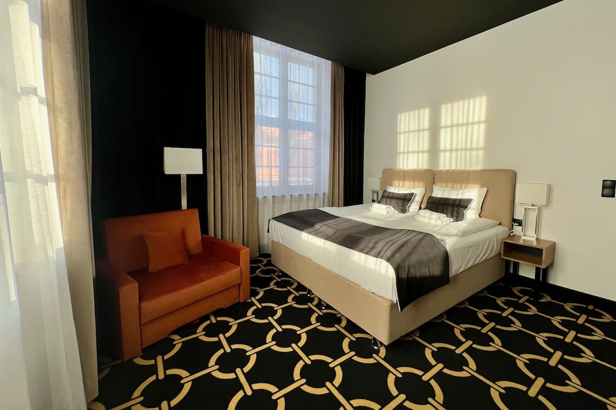 Quarto de hotel com uma cama grande, um sofá, uma mesinha de cabeceira e uma janela com cortinas.