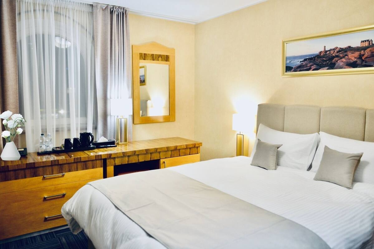 Quarto de hotel com cama, escrivaninha de madeira, espelho, mesa de cabeceira e um quadro na parede.