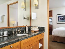 Casa de banho de hotel com bancada de granito, pia, espelho, porta-toalhas e uma cama no quarto adjacente.