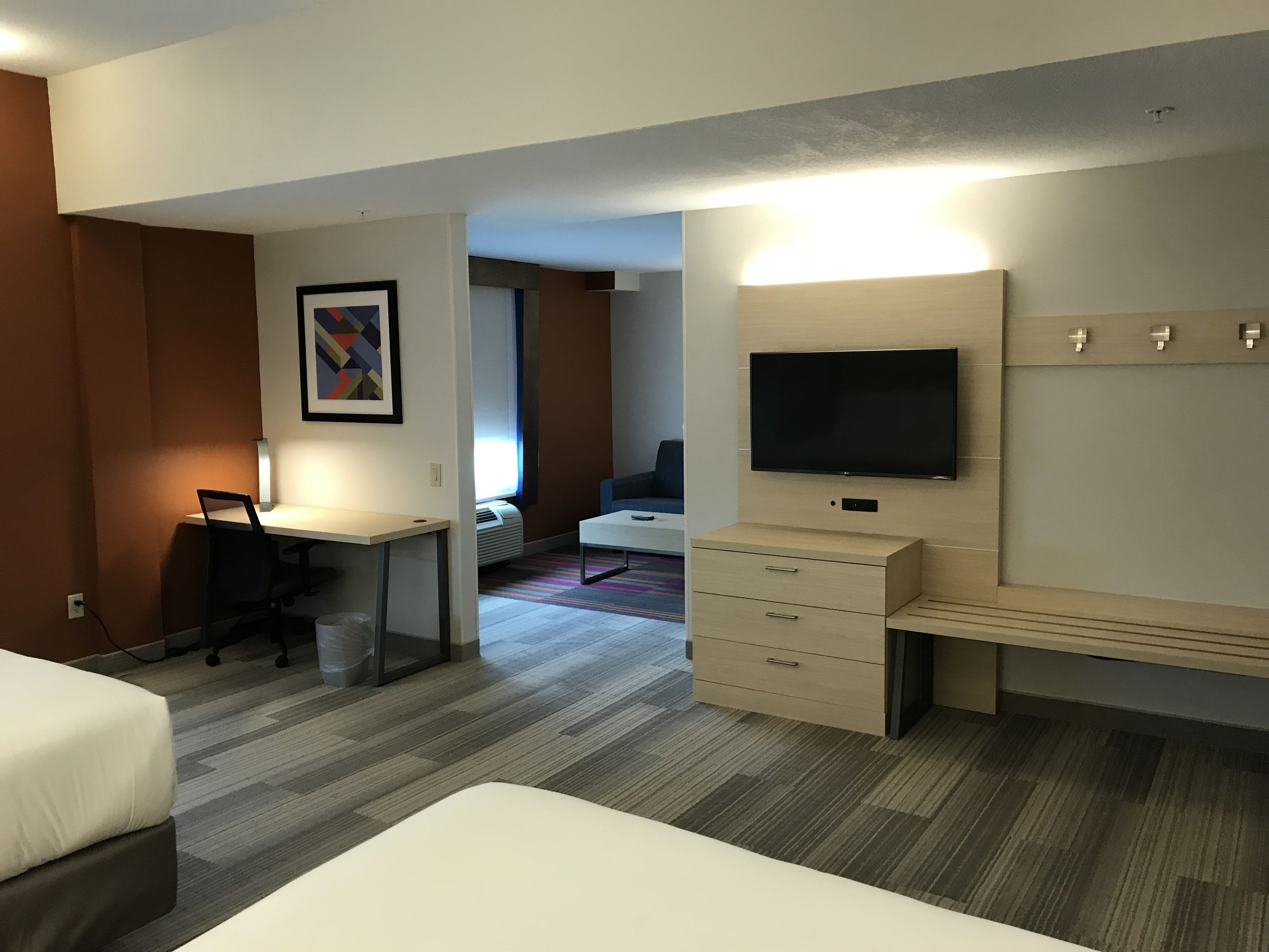 Quarto de hotel com cama, escrivaninha, cadeira, televisão e cómoda.