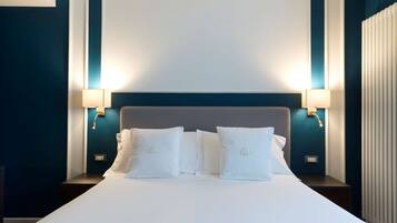 Cama bem arrumada com lençóis e almofadas brancas, com duas mesas de cabeceira com luminárias, em frente a uma parede azul escura com uma cabeceira grande e simples.
