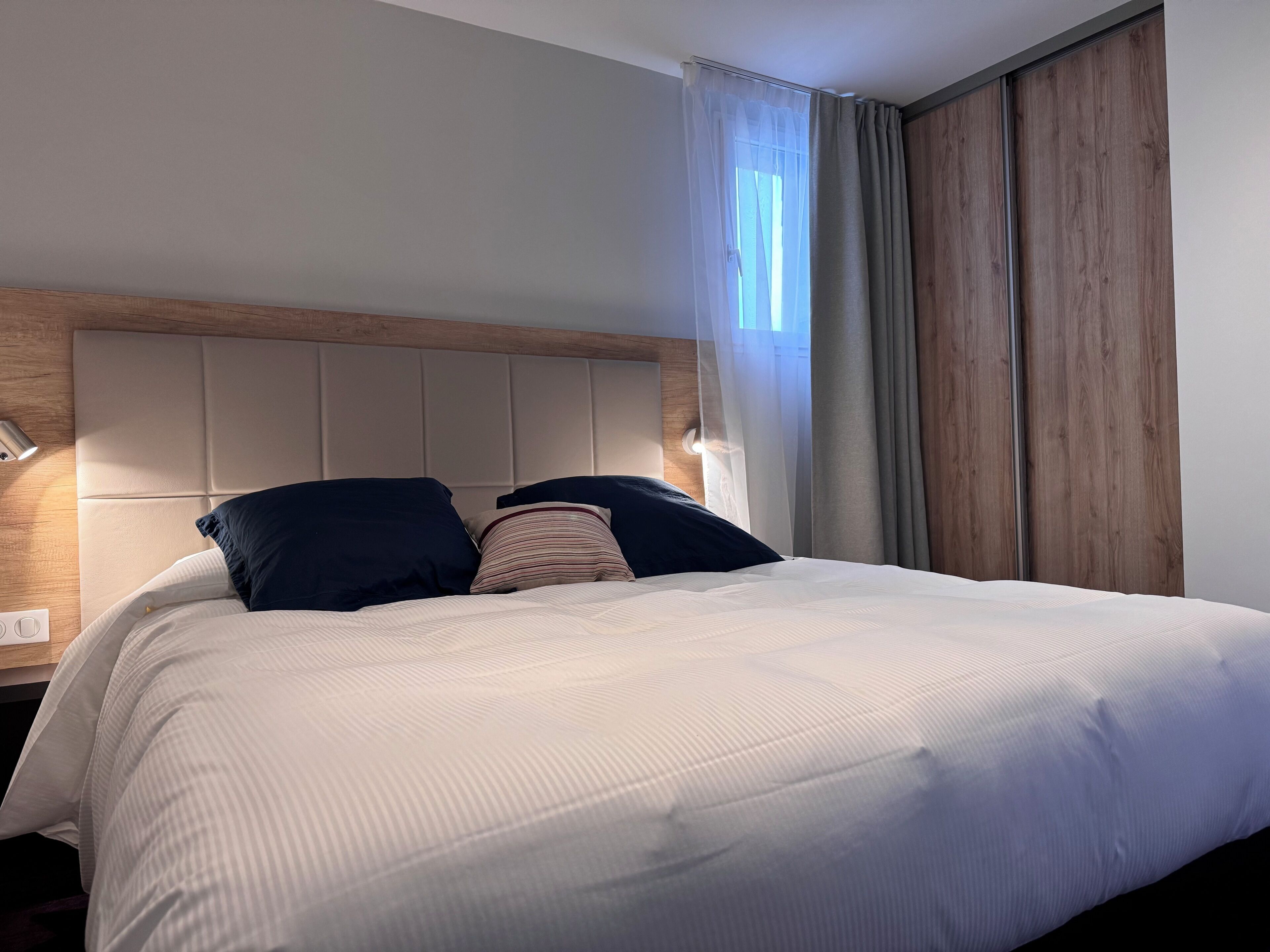 Uma cama bem arrumada com cabeceira, luminária de mesa e janela com cortinas leves.