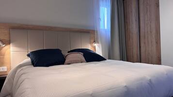 Uma cama bem arrumada com cabeceira, luminária de mesa e janela com cortinas leves.