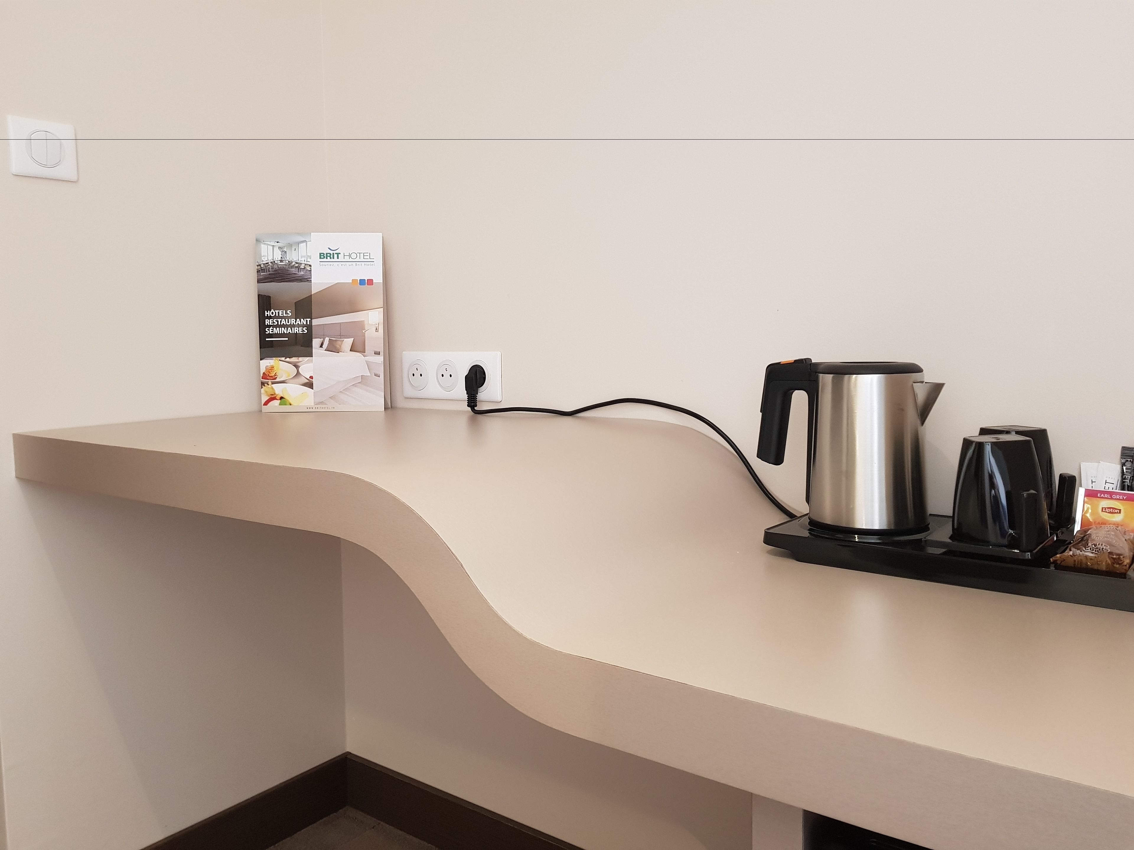 Uma área de recepção de hotel com um balcão curvo, chaleira, máquina de café e um folheto na parede.