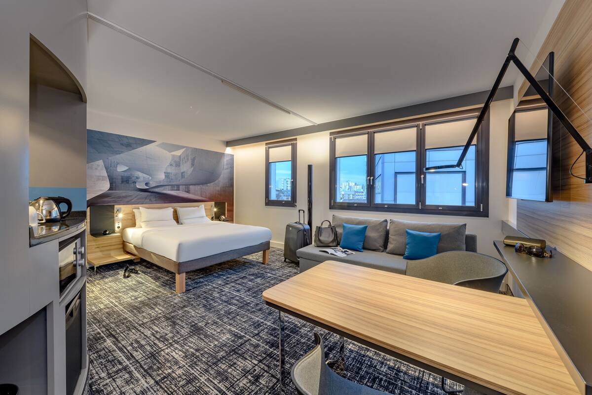 Quarto de hotel moderno com cama, sofá, escrivaninha e grandes janelas com vista para a cidade.