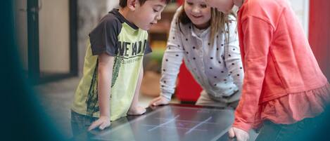 Duas pessoas interagindo com uma interface digital, possivelmente uma mesa touchscreen, em ambiente interno com parede vermelha e uma estante ao fundo.