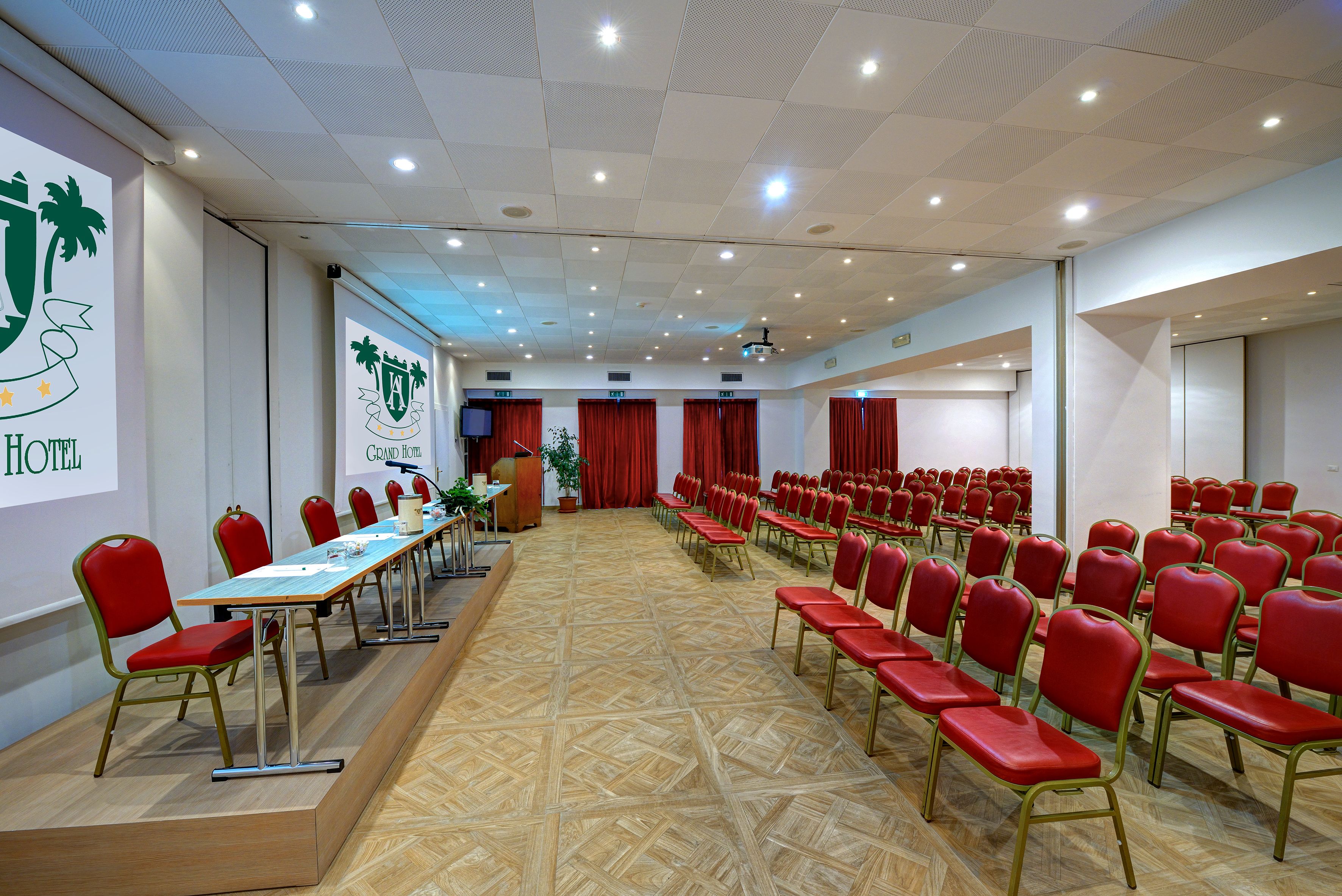Uma sala de conferência com fileiras de cadeiras vermelhas, uma mesa comprida com tela de projetor e piso estampado.
