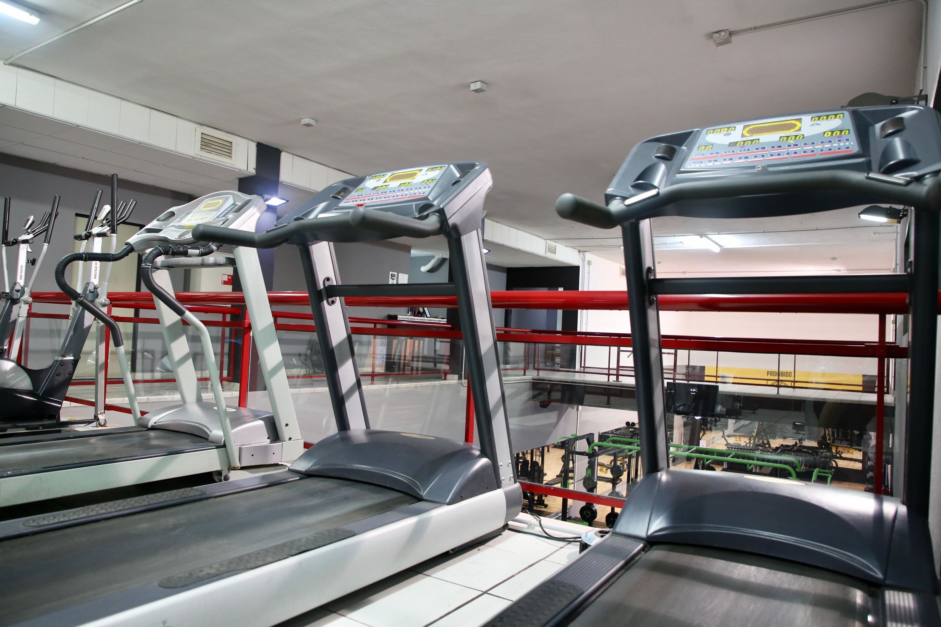 Uma academia com esteiras, equipamentos de ginástica e um corrimão vermelho.