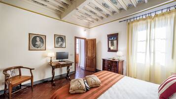 Quarto de hotel com uma cama, um banco de madeira, uma escrivaninha com abajur, uma televisão e dois retratos emoldurados na parede.