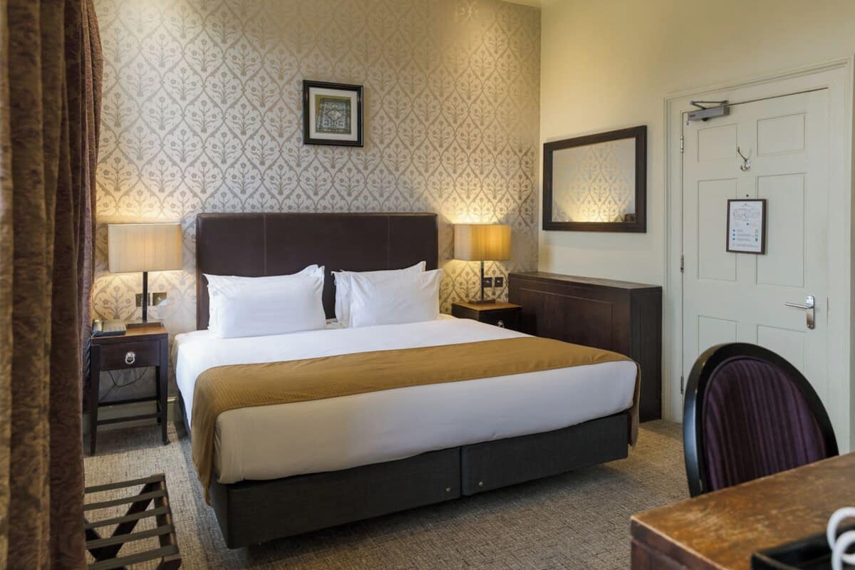 Quarto de hotel com uma cama grande, duas mesas de cabeceira, um criado-mudo, uma cadeira e uma porta com espelho.