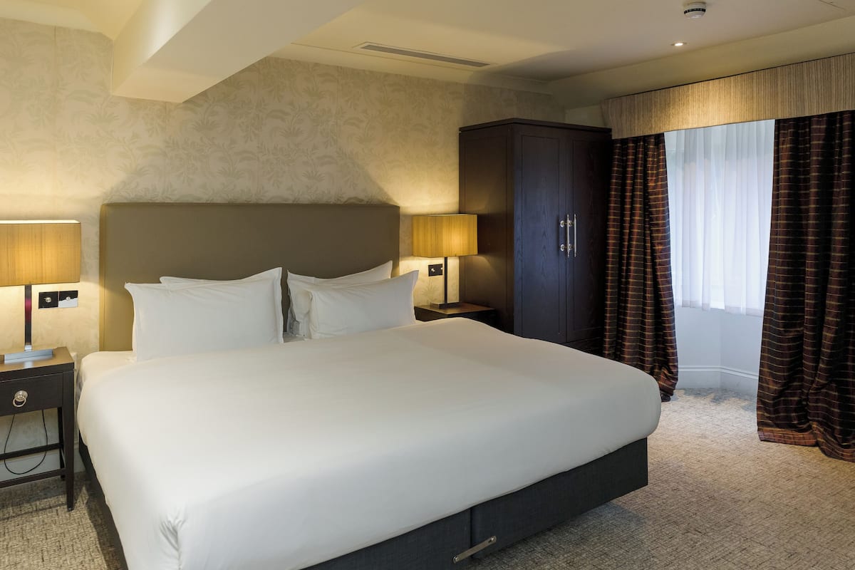 Quarto de hotel com uma cama grande, duas mesas de cabeceira com luminárias, um guarda-roupa e uma janela com cortinas.