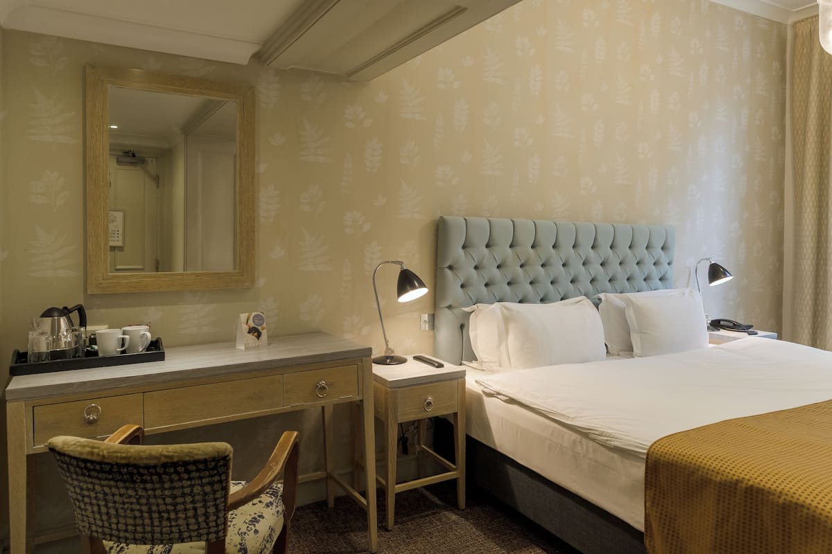 Quarto de hotel com cama, escrivaninha, cadeira, mesa de cabeceira, luminária e espelho.