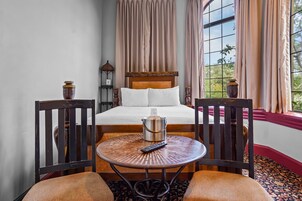 Quarto de hotel com cama, duas cadeiras, uma mesinha e janela com cortinas.