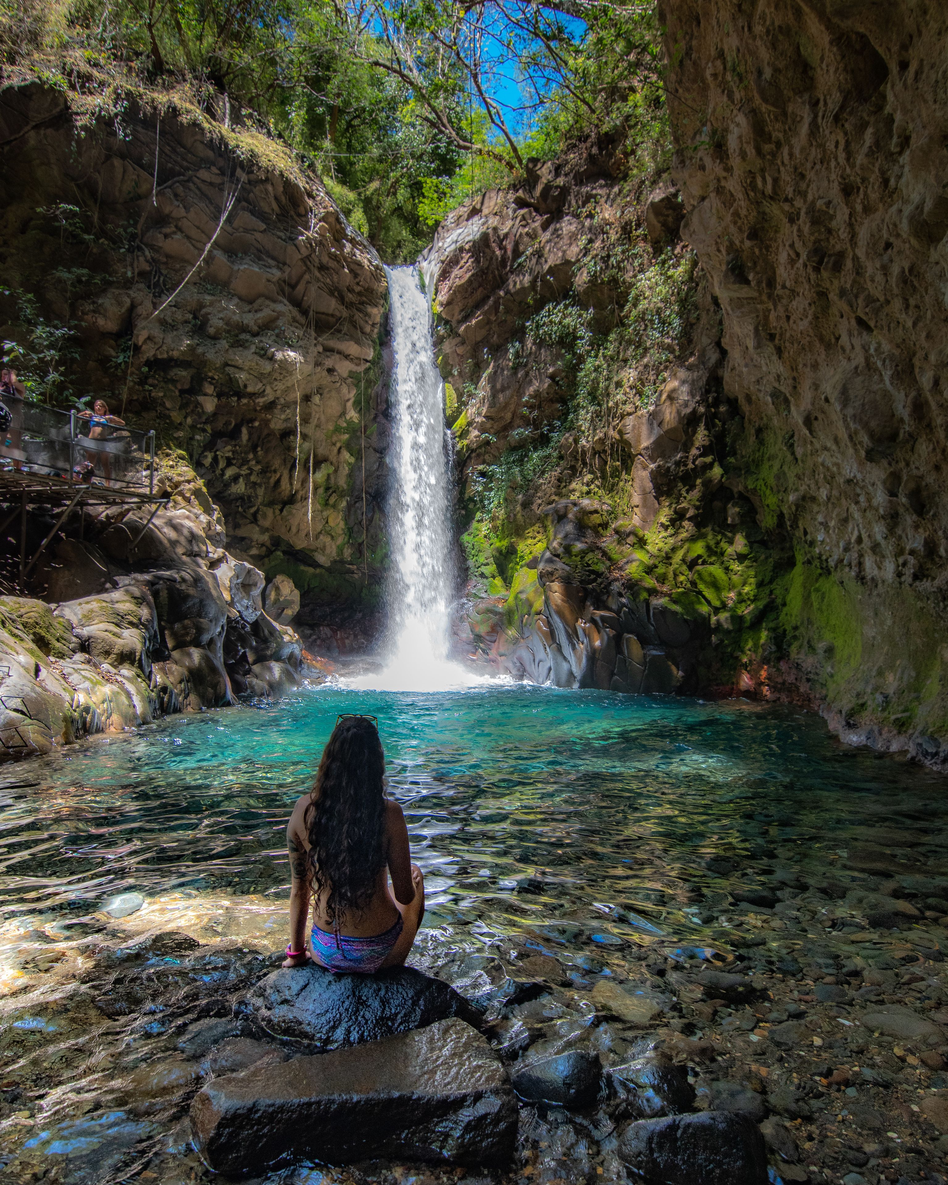 Uma cascata numa garganta rochosa com água cristalina e uma pessoa sentada sobre as pedras.
