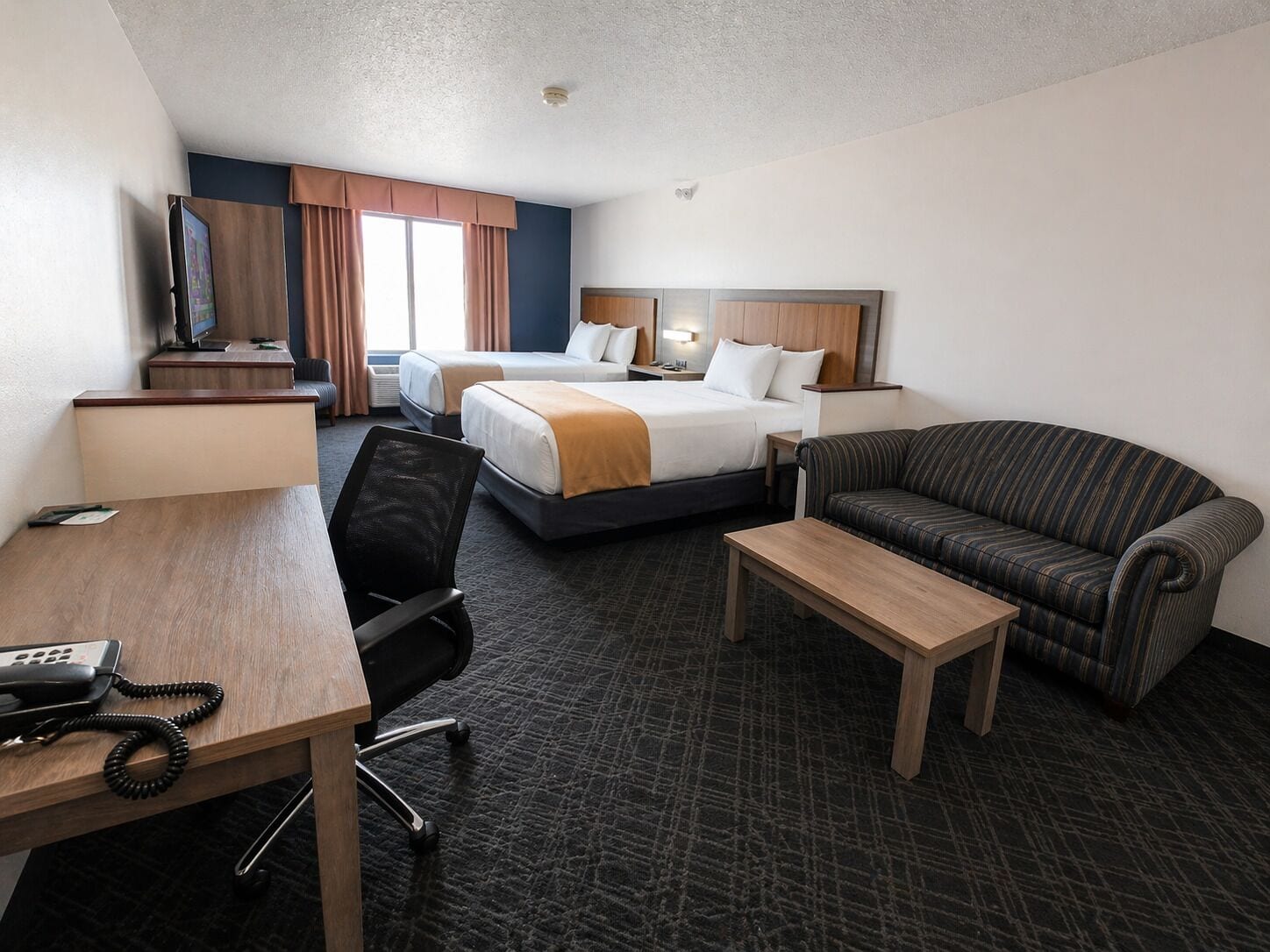 Quarto de hotel com duas camas, uma escrivaninha com telefone, uma cadeira, um sofá e uma televisão.