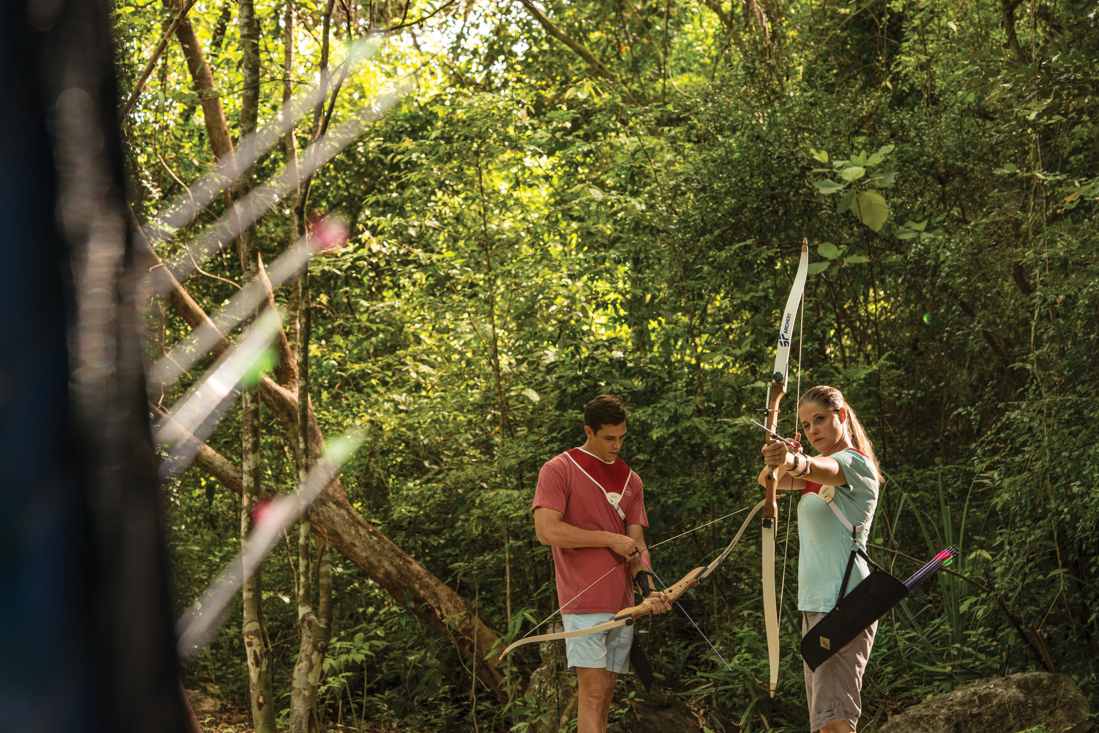 Duas pessoas praticando arco e flecha em uma área arborizada.