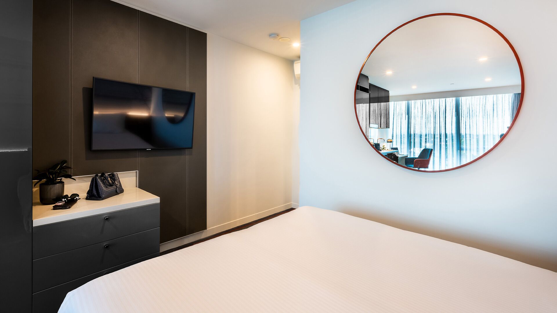 Quarto moderno com cama grande, televisão fixada na parede, mesa de cabeceira com bolsa e espelho redondo.