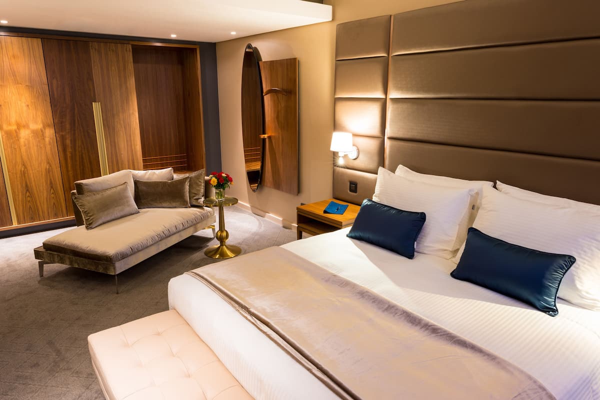 Quarto de hotel com uma cama grande, um sofá, uma mesa de cabeceira com abajur e um guarda-roupa de madeira.