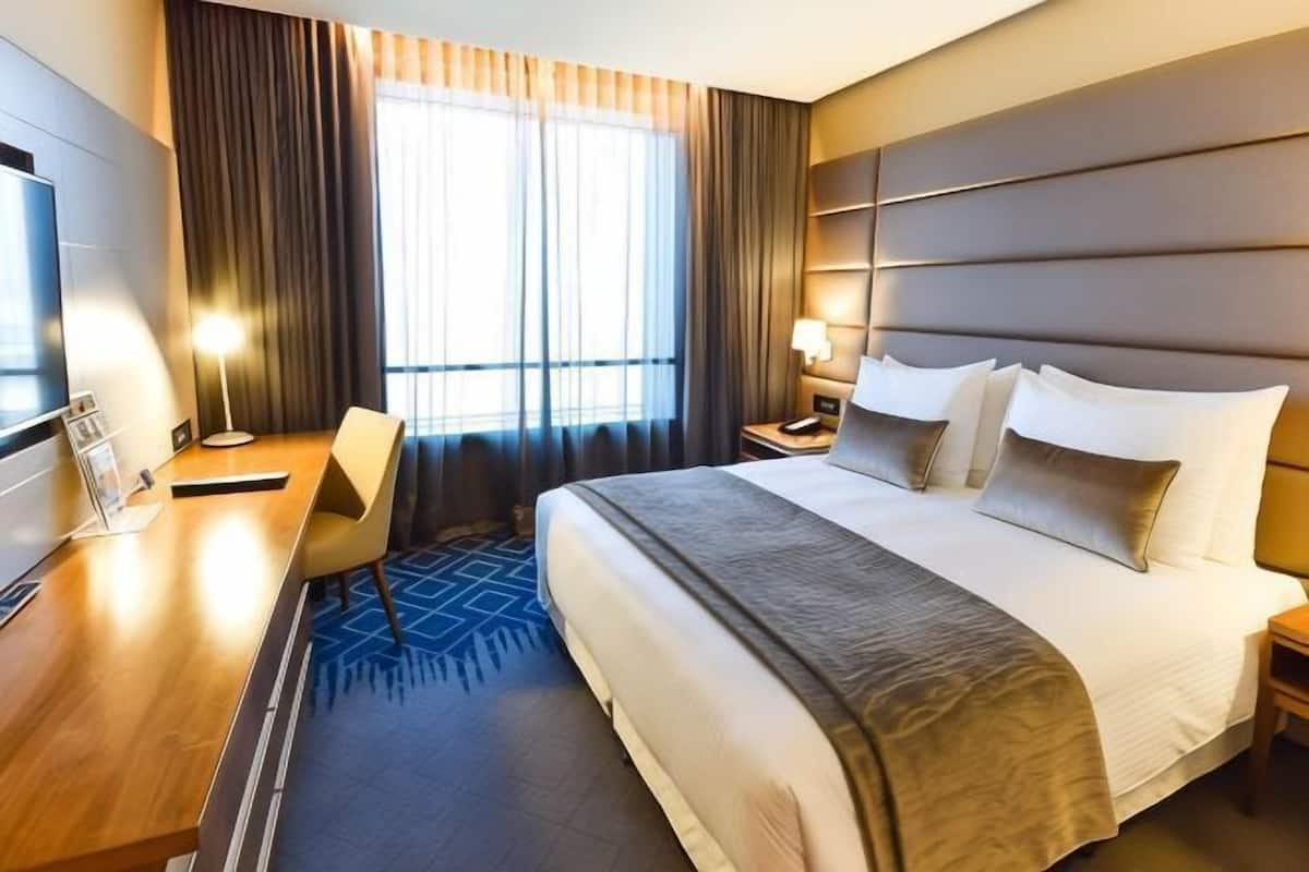 Quarto de hotel com uma cama grande, uma escrivaninha, uma cadeira, uma televisão e uma janela com cortinas.