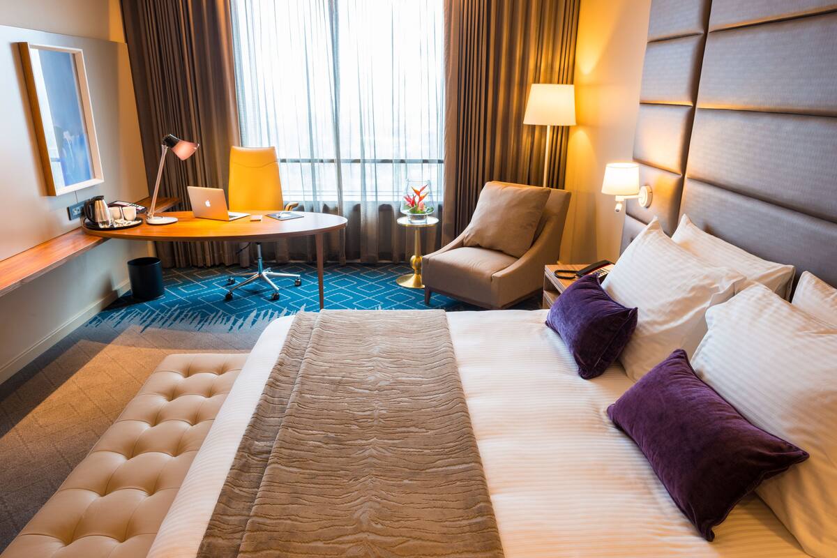 Quarto de hotel com uma cama grande, uma escrivaninha com um laptop, uma cadeira e uma pequena mesa com abajur.
