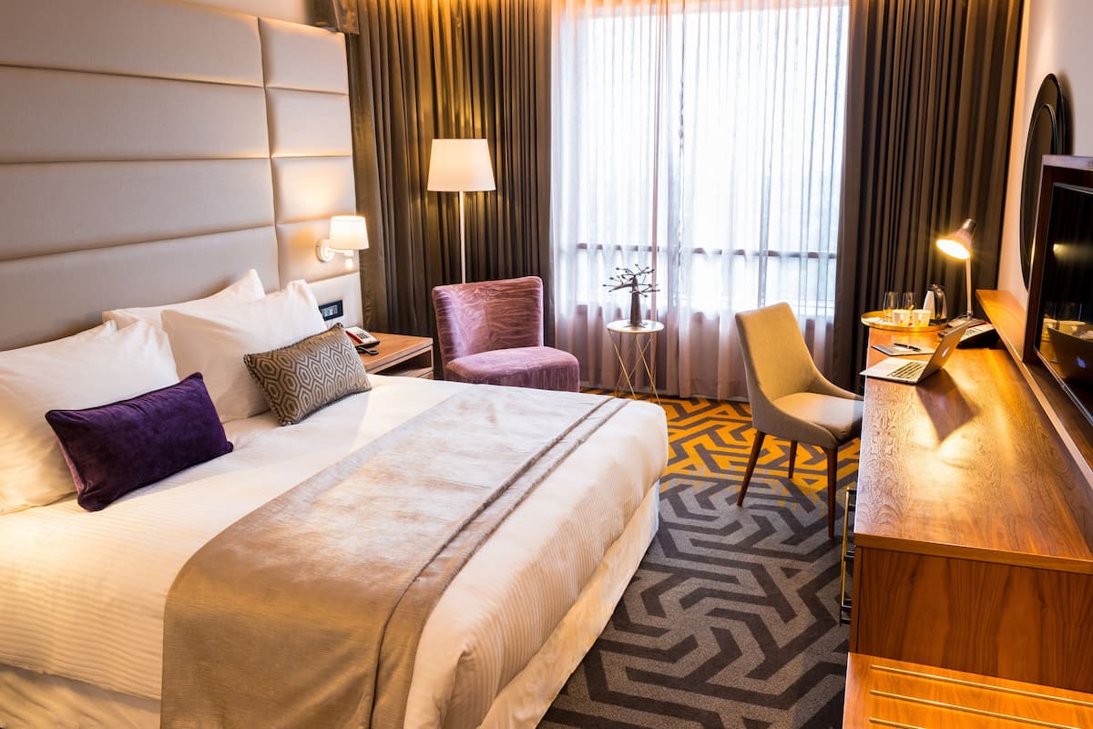 Quarto de hotel com uma cama grande, uma escrivaninha com um laptop, uma cadeira, uma pequena mesa com abajur e uma TV.