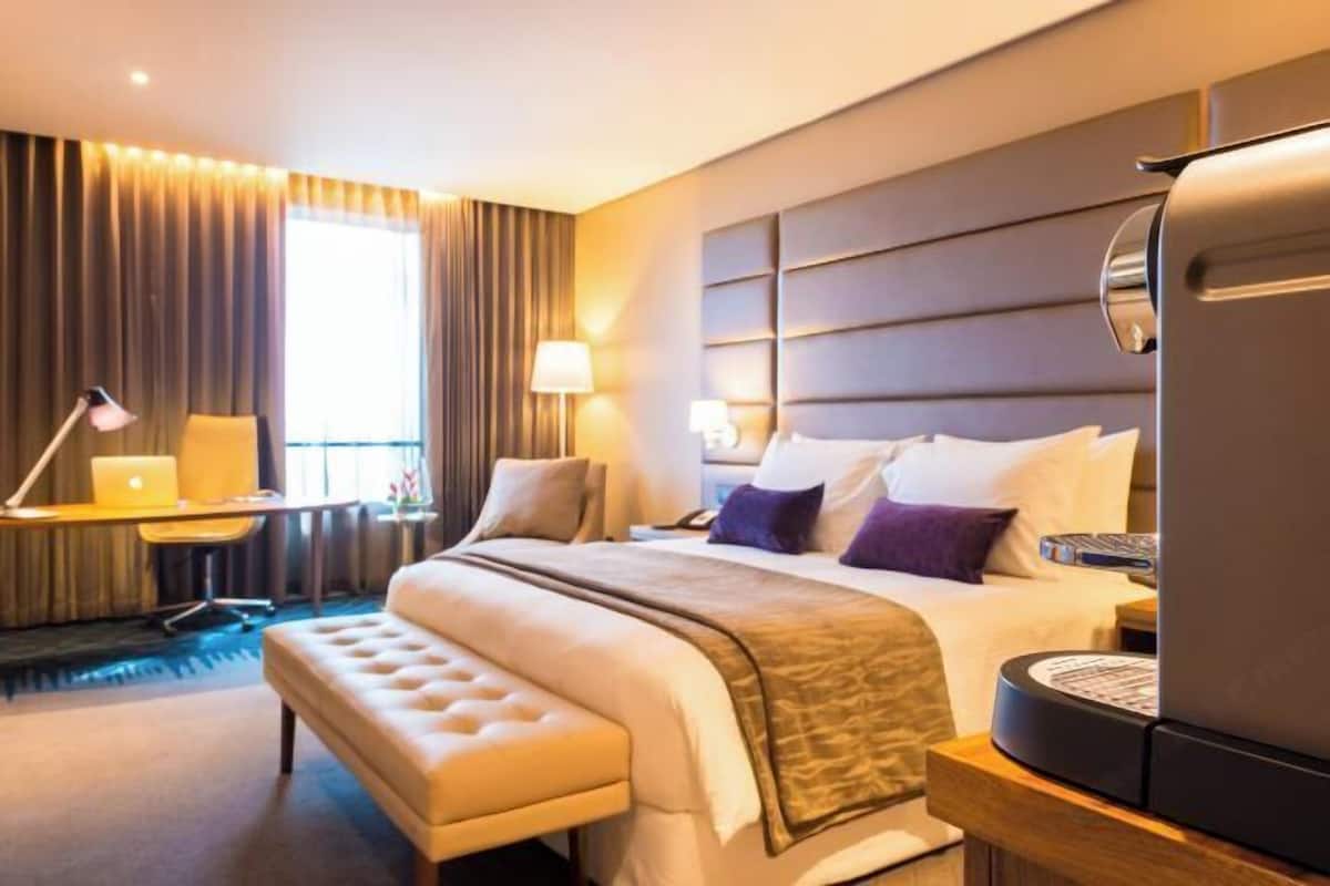 Quarto de hotel com cama, mesas de cabeceira, escrivaninha com um laptop, uma cadeira e uma janela grande com cortinas.