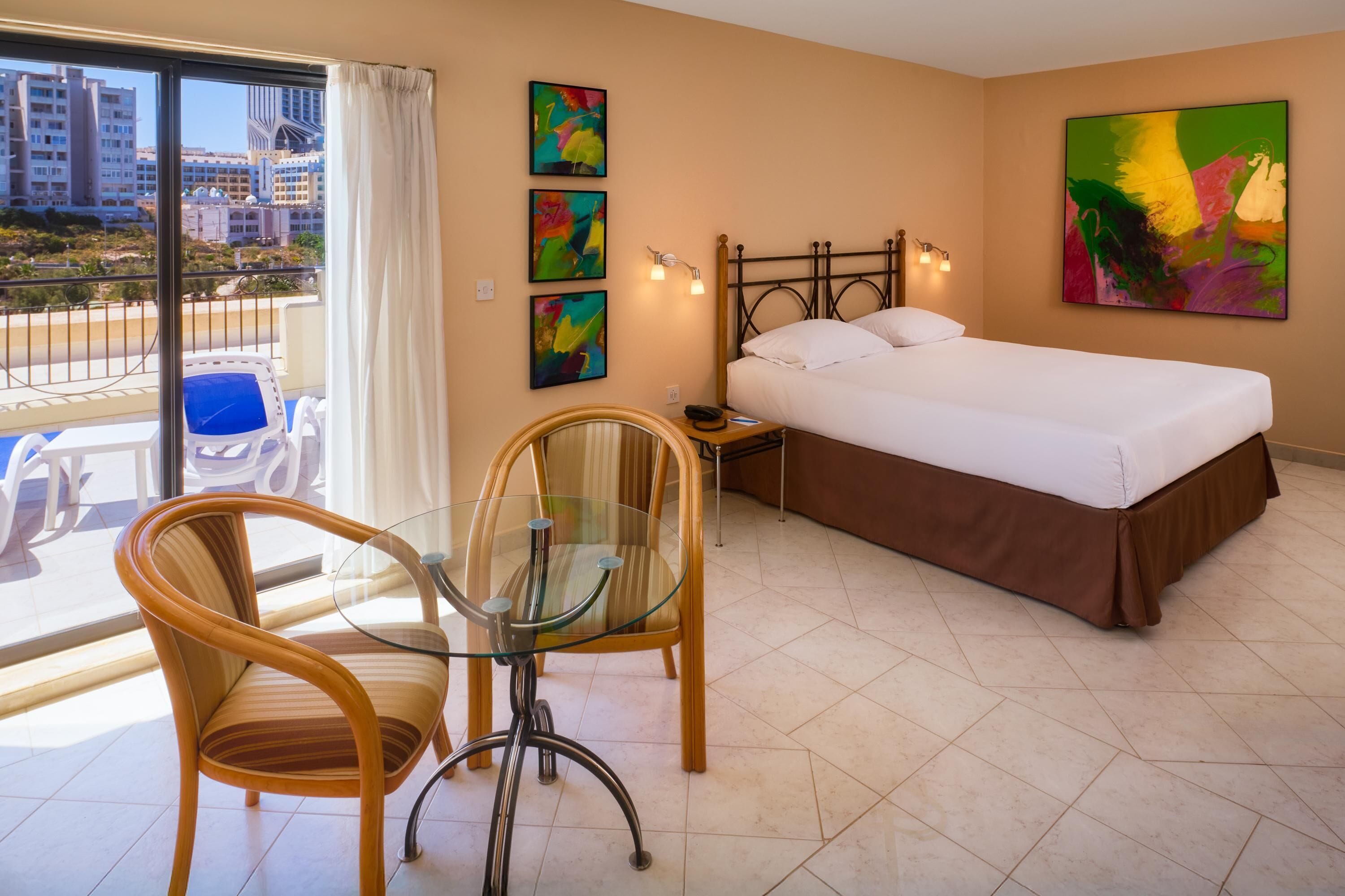 Quarto de hotel com uma cama, mesa de vidro com cadeiras, varanda com vista para edifícios e três quadros abstratos na parede.