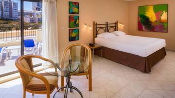 Quarto de hotel com uma cama, mesa de vidro com cadeiras, varanda com vista para edifícios e três quadros abstratos na parede.