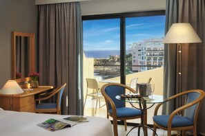 Quarto de hotel com varanda, cama, escrivaninha com abajur, cadeira e mesa com tampo de vidro.