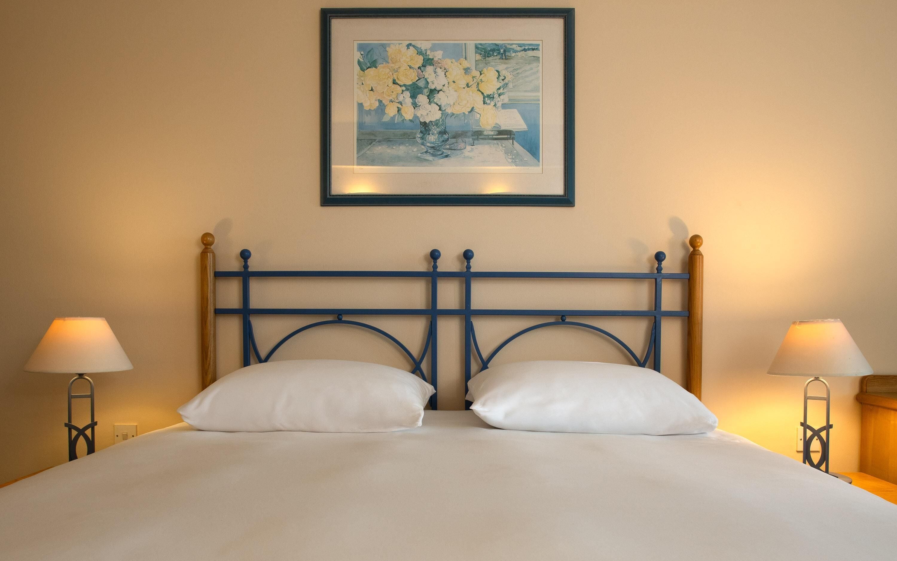 Uma cama com lençóis brancos, cabeceira de metal azul, duas luminárias de cabeceira e um quadro emoldurado sobre a cama.