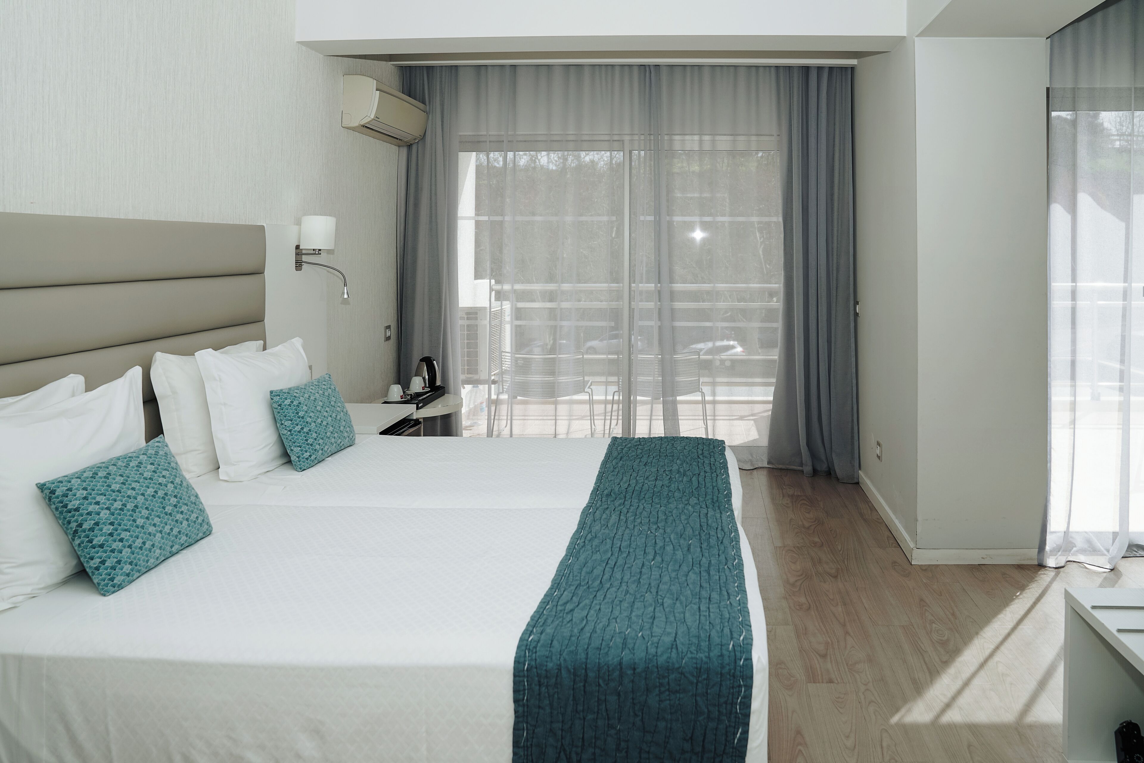 Quarto de hotel com uma cama grande, uma mesa de cabeceira, uma luminária e vista para o exterior através de uma janela ampla.
