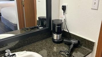 Casa de banho de hotel com lavatório branco, espelho, máquina de café e secador de cabelo.